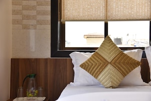 Quarto de hotel com cama, janela com vista para edifícios, cabeceira de madeira e mesa de cabeceira com um pote de vidro e uma garrafa verde.