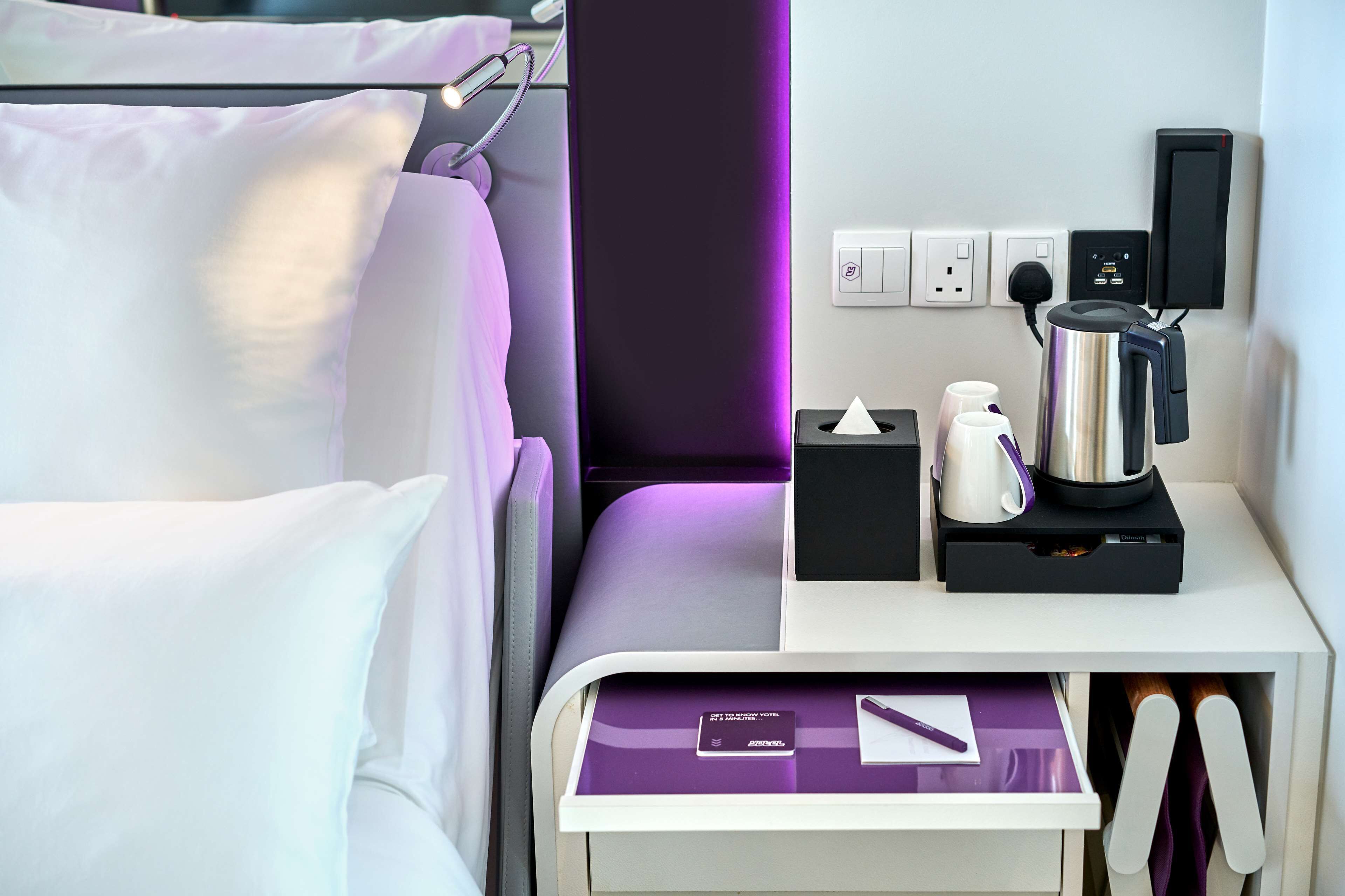 Quarto de hotel com cama, mesa de cabeceira com cafeteira, caixa de lenços e mesinha com caneta e cartão.