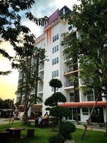 Um hotel com vários andares, fachada branca, varandas e uma pequena área de jardim com bancos.
