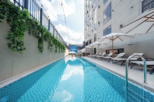 Piscina com espreguiçadeiras e guarda-sóis, cercada por um edifício com fachada em azulejos e vegetação.