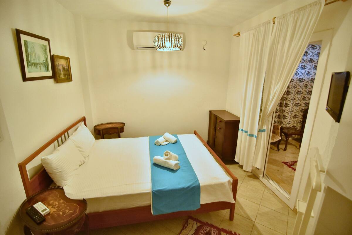 Cama bem arrumada com colcha azul e duas toalhas enroladas, mesa de cabeceira de madeira com um controle remoto, pequena mesa de madeira, ar-condicionado fixado na parede e vista através de uma cortina para um quarto com padrão.