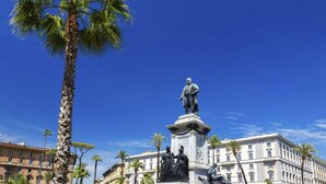 Uma estátua em frente a um edifício com arquitetura clássica, palmeiras e céu azul claro.