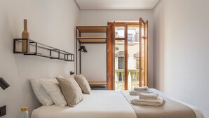 Uma cama bem arrumada com roupa de cama branca e bege, uma mesa de cabeceira com uma garrafa e um porta-toalhas.