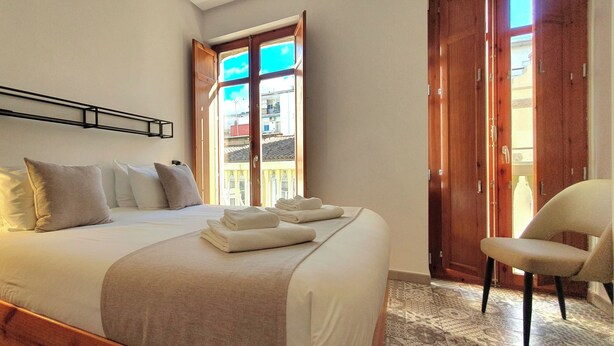 Cama bem arrumada com almofadas e toalhas, uma mesa de cabeceira, uma cadeira e uma janela com vista para prédios.