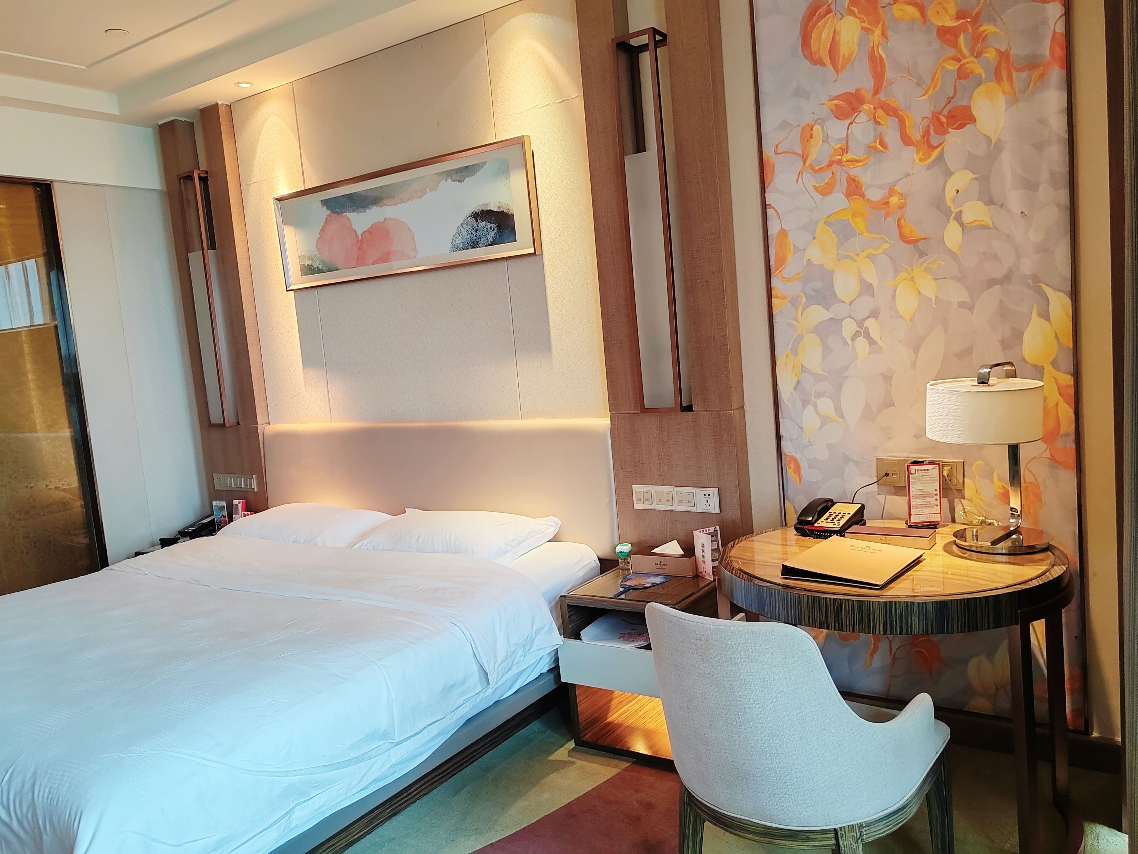 Quarto de hotel com uma cama, escrivaninha com abajur, uma cadeira e um quadro na parede.