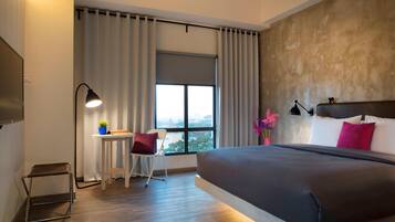 Quarto de hotel moderno com uma cama grande, uma escrivaninha com cadeira, uma janela com cortinas e vista para a cidade.