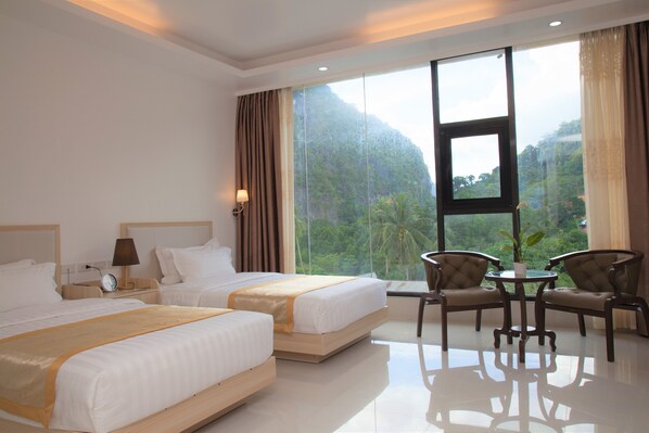 Quarto de hotel com duas camas, uma mesinha, duas cadeiras, janela com vista para uma montanha e vegetação, e uma TV fixada na parede.