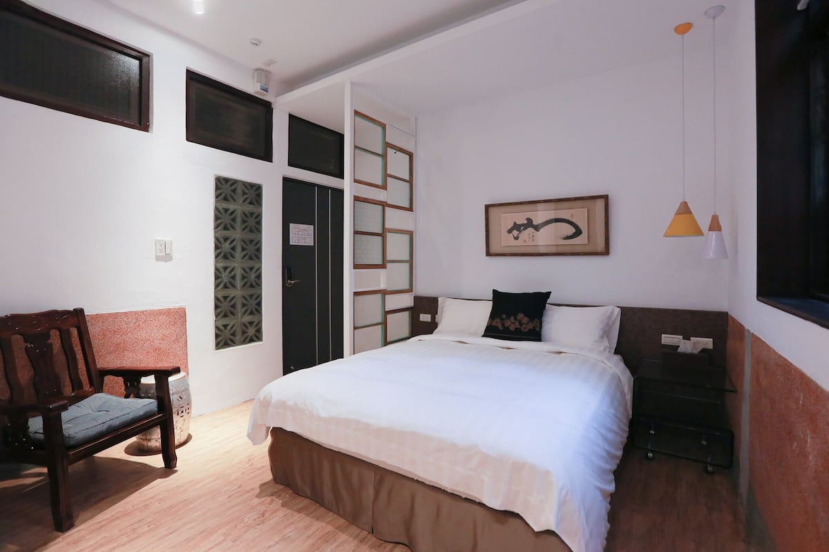 Quarto com cama, cadeira, mesa de cabeceira e prateleira na parede.