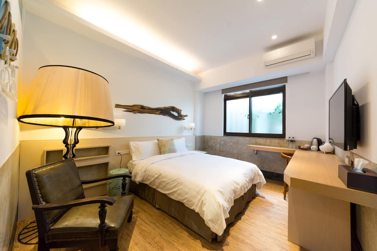 Quarto de hotel moderno com uma cama grande, uma escrivaninha, uma cadeira, um abajur e uma televisão.
