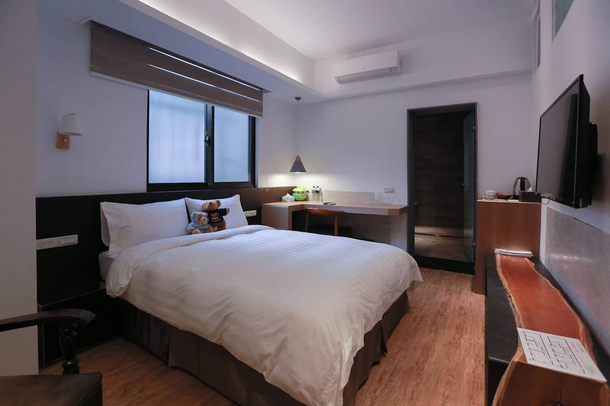 Quarto de hotel com uma cama grande, mesa de cabeceira, luminária, TV e escrivaninha com cadeira.