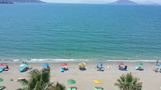 Uma praia com vários guarda-sóis e espreguiçadeiras, vista clara para o mar e montanhas ao longe.