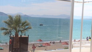 Vista da praia por uma janela, com uma palmeira, guarda-sóis e uma torre de salva-vidas.