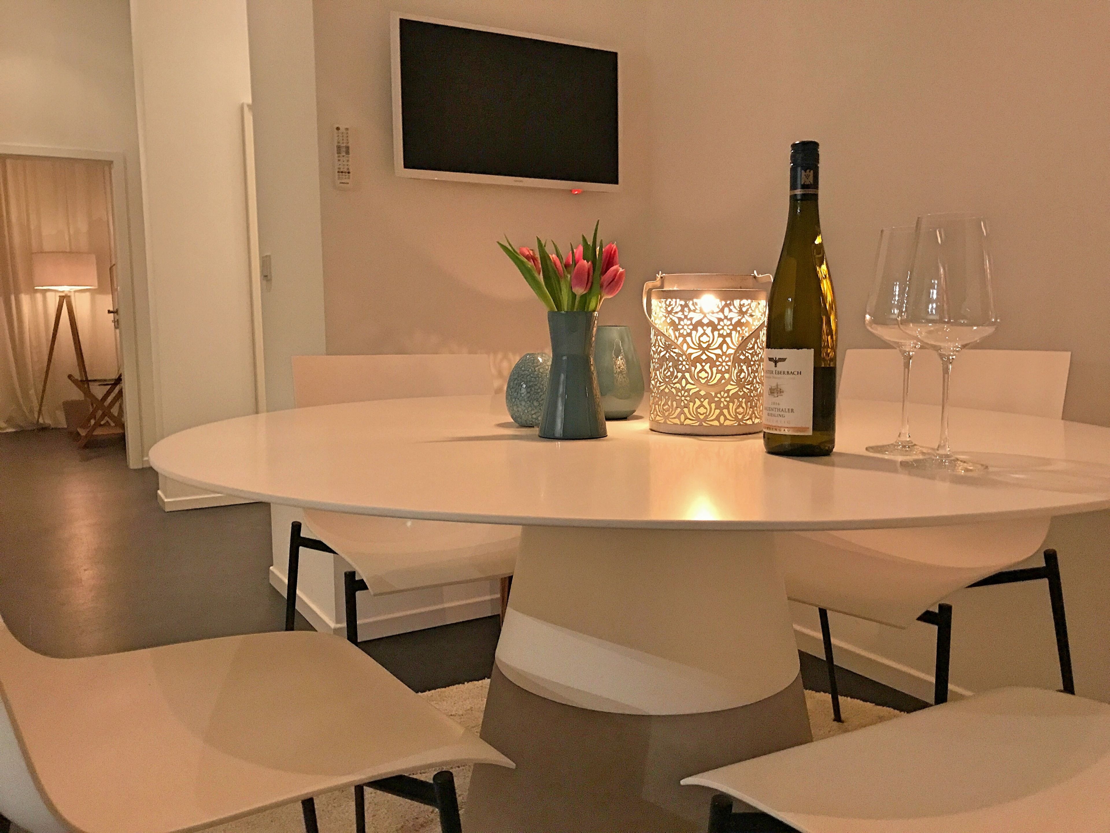 Uma mesa de jantar redonda com cadeiras, uma garrafa de vinho, duas taças de vinho, um vaso com flores e um castiçal decorativo.