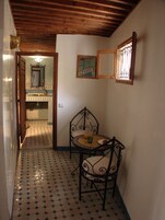 Um corredor estreito com chão de azulejos que dá para um banheiro. Há uma mesa com uma cadeira no corredor e dois espelhos nas paredes.