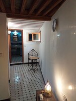 Um corredor estreito com chão de azulejos, uma pequena mesa com uma cadeira, um relógio na parede e um abajur aceso.