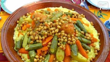 Uma grande tigela redonda e marrom cheia de cuscuz, grão de bico, cenouras, abobrinha e outros vegetais.