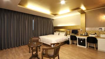 Quarto de hotel com cama, escrivaninha, cadeiras, uma pequena mesa, janela e cozinha compacta.