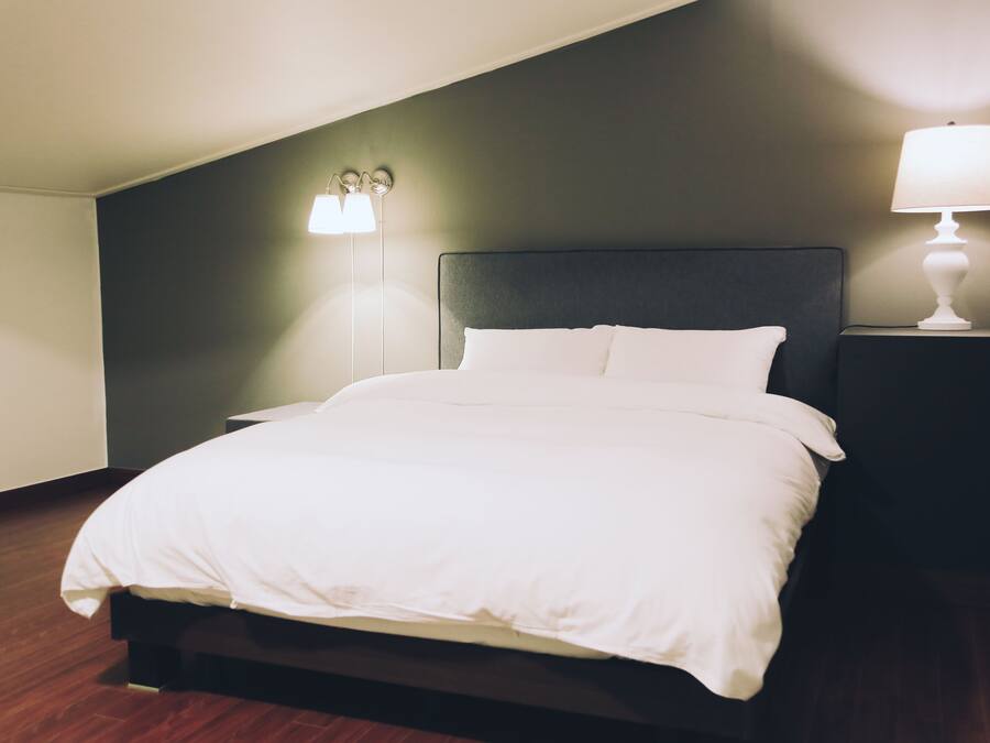 Cama bem arrumada com roupa de cama branca em quarto de hotel moderno, com cabeceira escura e mesa de cabeceira com abajur.