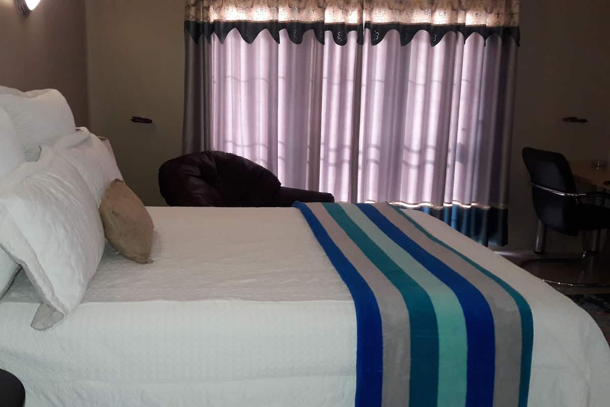 Quarto de hotel com uma cama grande, almofadas, manta listrada, uma cadeira, escrivaninha e janela com cortinas.