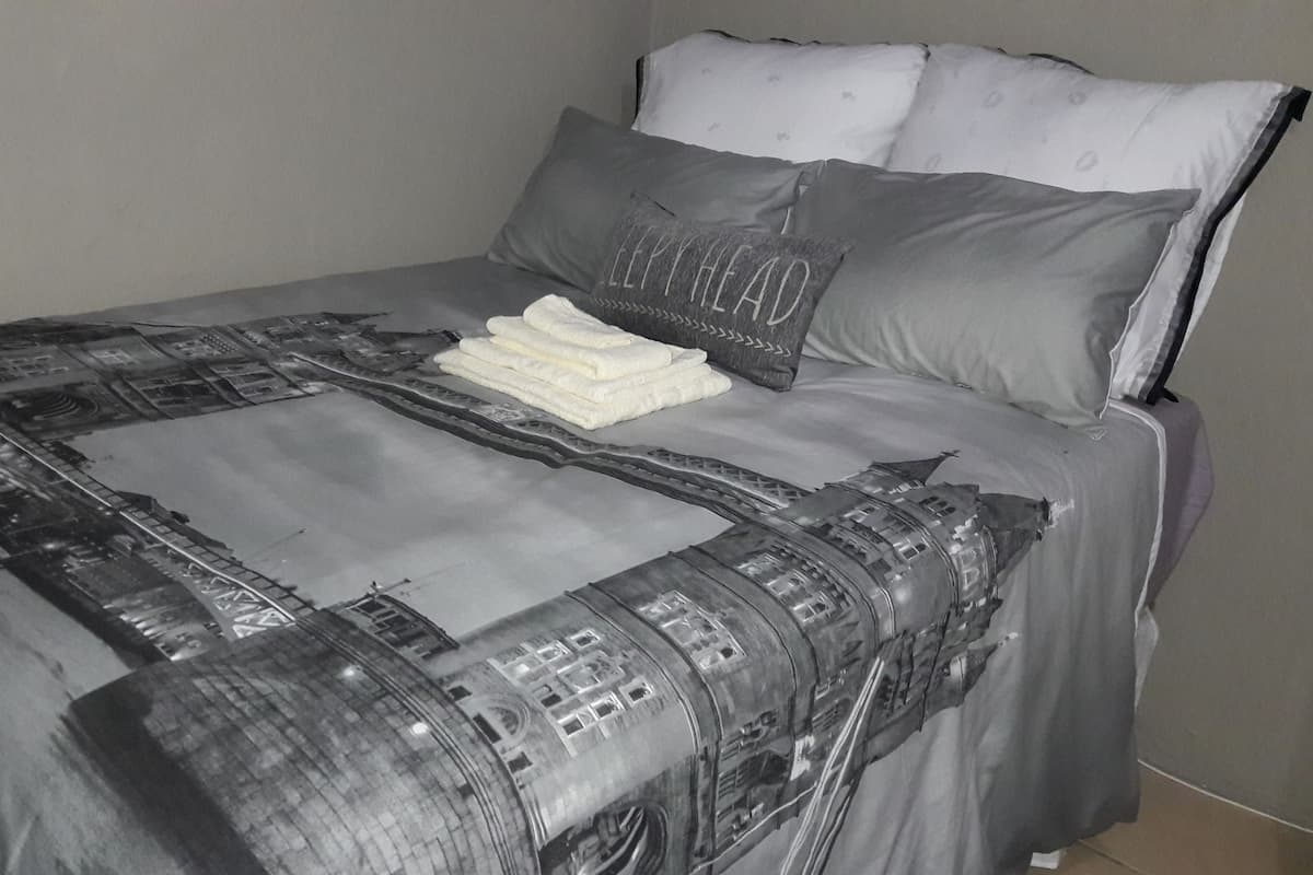 Uma cama com colcha cinzenta e almofadas, uma toalha branca dobrada e uma almofada decorativa estampada.