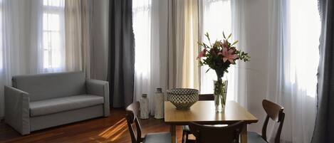 Uma área de refeições com uma mesa de madeira, duas cadeiras, um vaso com flores e um sofá cinzento.