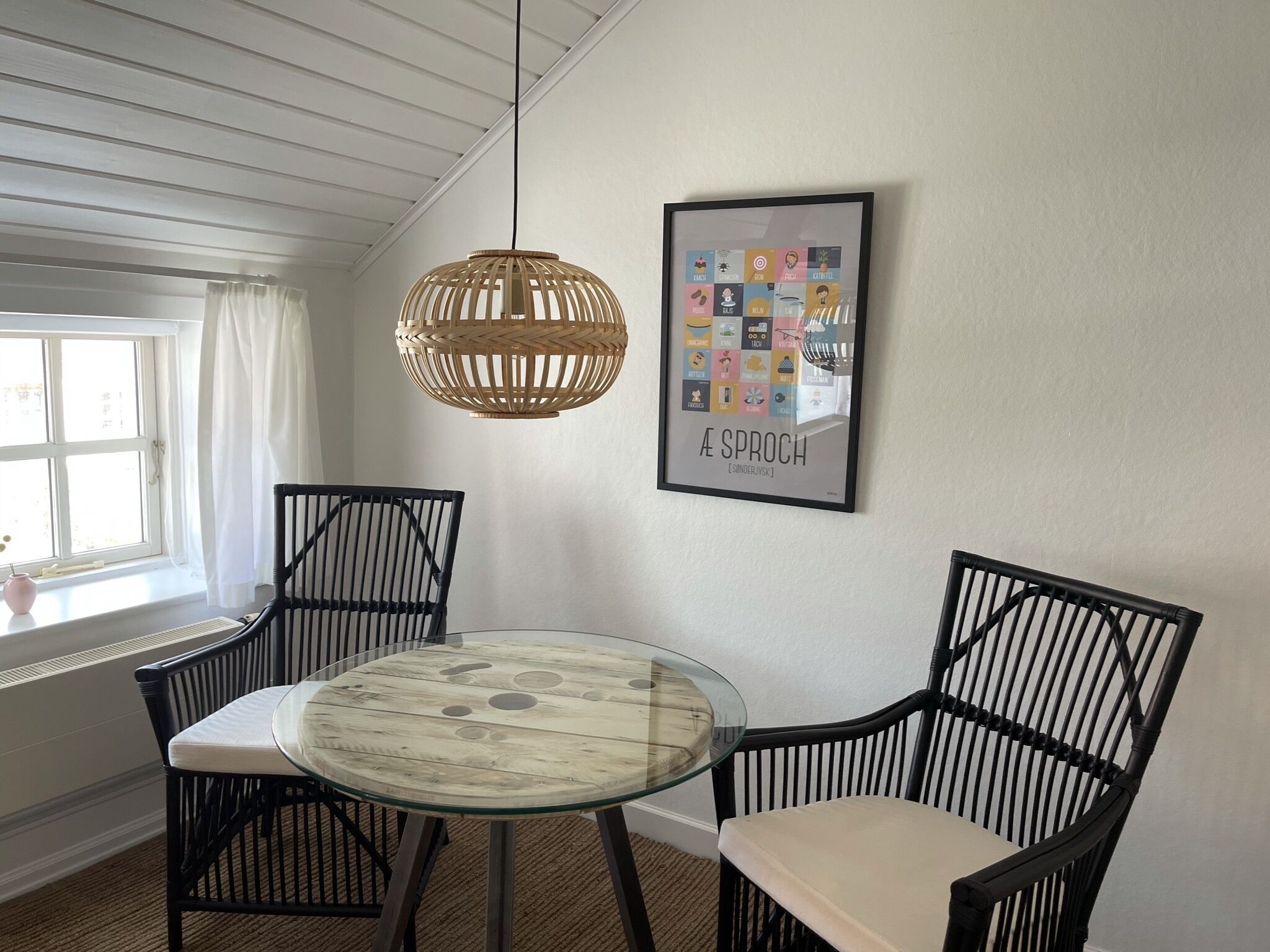 Uma área de jantar com uma mesa redonda de vidro, cadeiras pretas, uma luminária pendente trançada e um pôster emoldurado na parede.