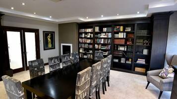 Um quarto espaçoso com uma grande mesa de madeira escura rodeada por cadeiras, uma estante cheia de livros e uma poltrona cinza com uma almofada colorida.