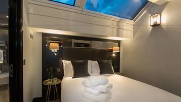 Quarto de hotel com claraboia, cama com roupa de cama branca, mesa de cabeceira pequena com abajur e luminária na parede.
