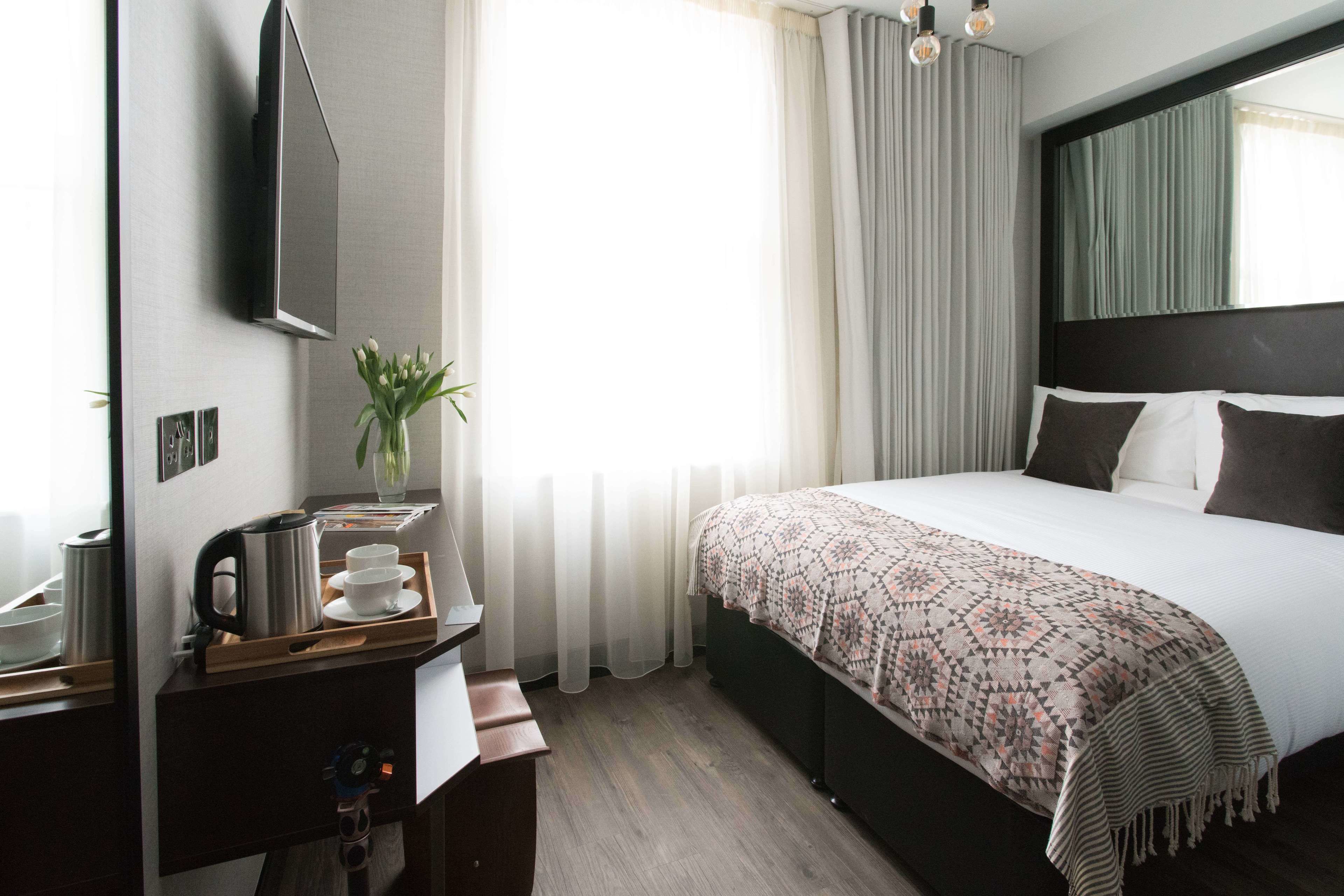 Quarto de hotel moderno com uma cama bem arrumada, uma mesinha de cabeceira com uma bandeja de itens de café da manhã, um espelho grande e uma janela com cortinas.