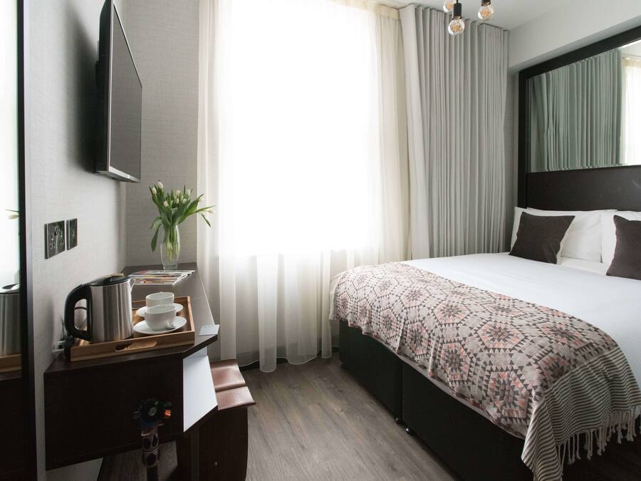 Quarto de hotel moderno com uma cama bem arrumada, uma mesinha de cabeceira com uma bandeja de itens de café da manhã, um espelho grande e uma janela com cortinas.