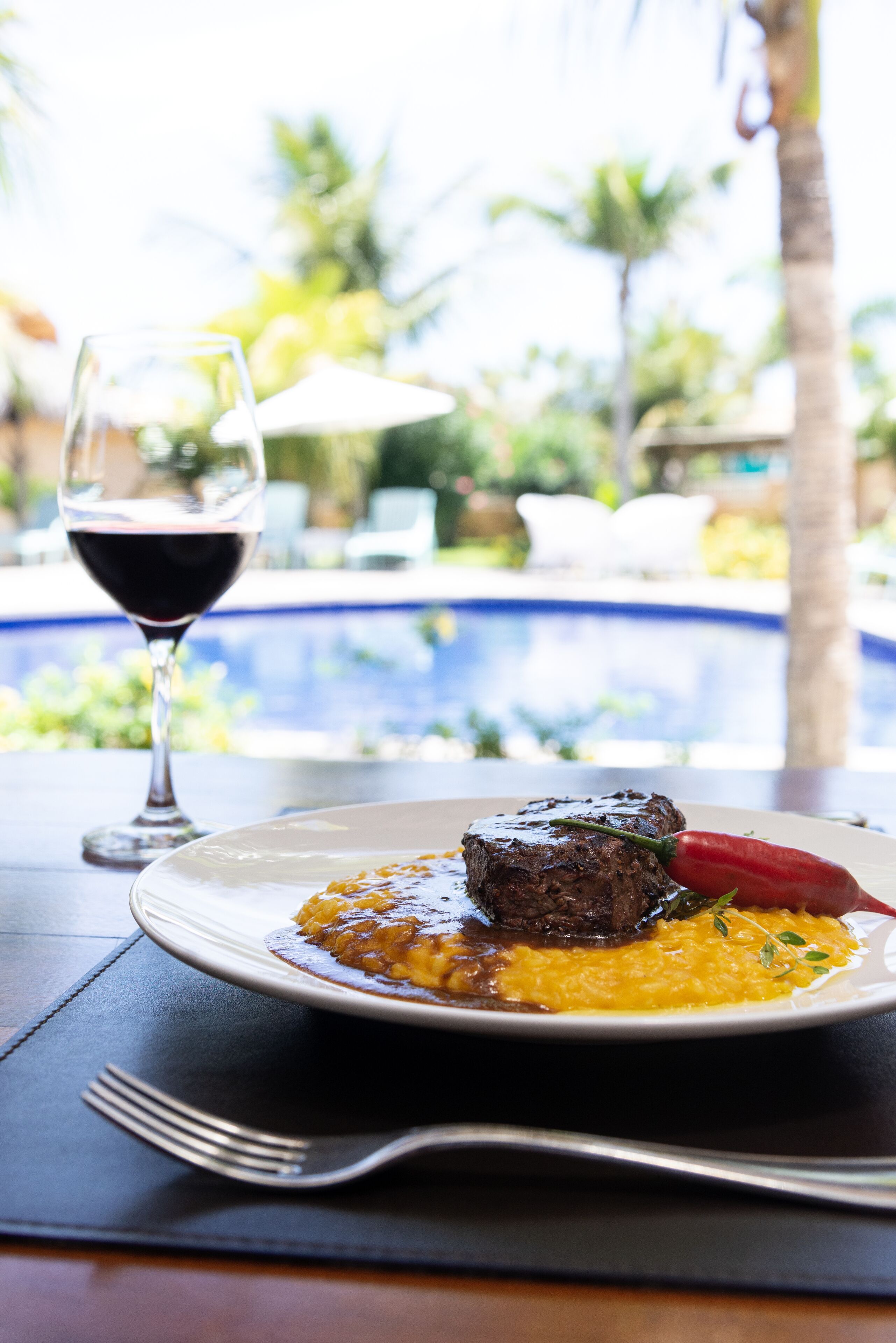 Uma mesa de jantar com um prato de comida, um copo de vinho tinto e uma piscina ao fundo.