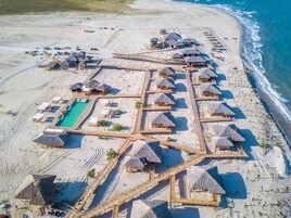 Vista aérea de um resort à beira-mar com chalés de telhado de palha dispostos em padrão de grade, uma piscina central e uma praia arenosa.