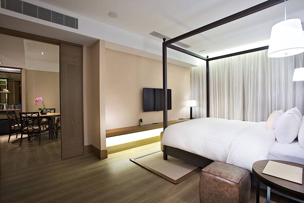 Quarto de hotel com cama de dossel, uma escrivaninha com cadeira, televisão de tela plana e área de jantar com cadeiras e mesa.