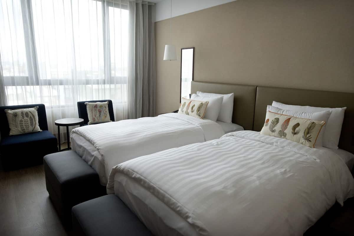 Quarto de hotel com duas camas, uma mesa redonda pequena, um sofá e uma janela grande com cortinas leves.