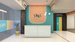 Uma área de recepção com design moderno, com um balcão de atendimento, o logotipo da Sans Hotels e a placa do hotel.