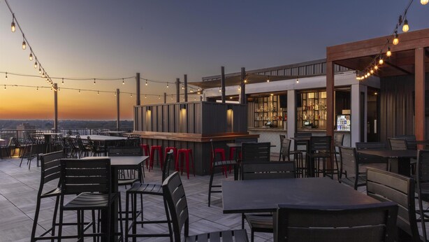 Uma área de refeições ao ar livre com mesas, cadeiras, luzes de Natal e um bar com vista para o horizonte da cidade.