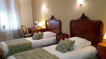 Quarto de hotel com duas camas, cada uma com roupa de cama estampada com flores, cabeceiras de madeira, mesas de cabeceira com abajures e uma janela com cortinas finas.