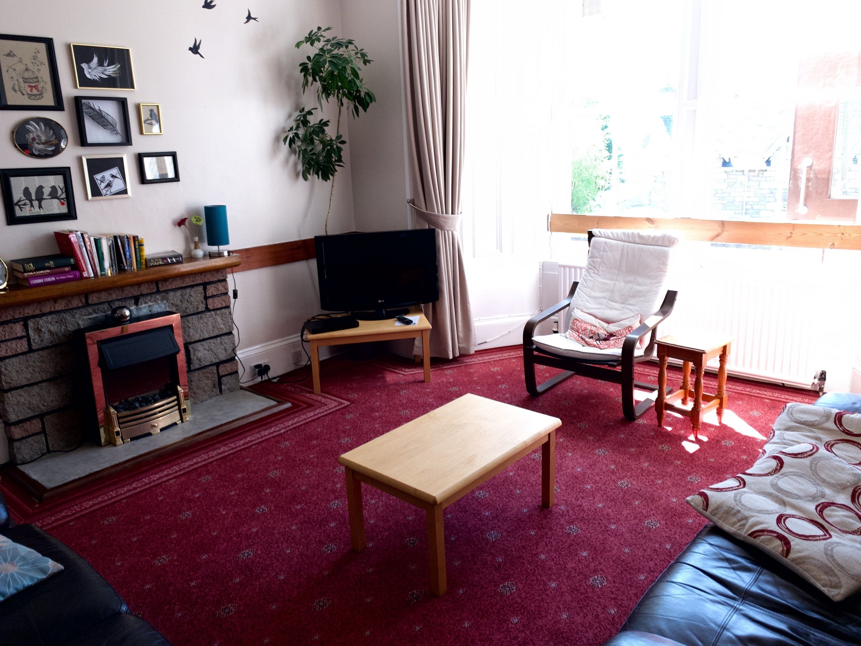 Uma sala de estar com lareira, televisão, tapete vermelho, mesa de centro de madeira, sofá azul e uma janela com cortinas.