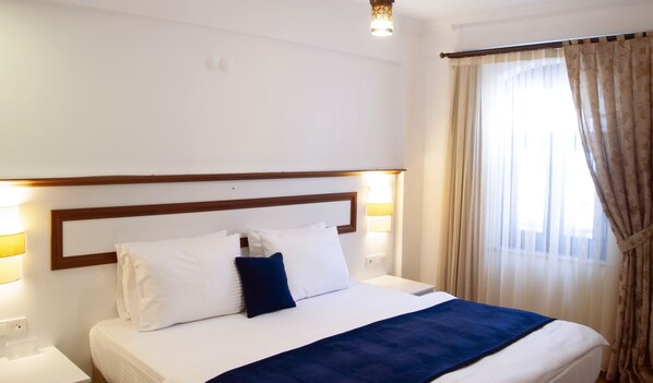 Quarto de hotel com uma cama grande, cabeceira de madeira, duas luminárias de apoio e uma janela com cortinas.