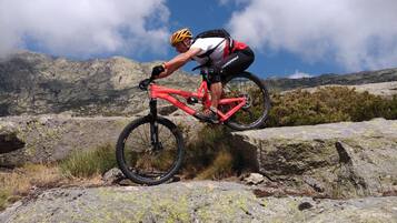 Um ciclista de montanha com uma bicicleta vermelha e uma mochila, pedalando em terreno rochoso com montanhas ao fundo.