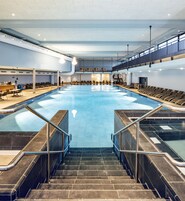 Uma piscina interior ampla com superfície de água azul límpida, rodeada por espreguiçadeiras e uma escadaria que dá para um segundo andar.