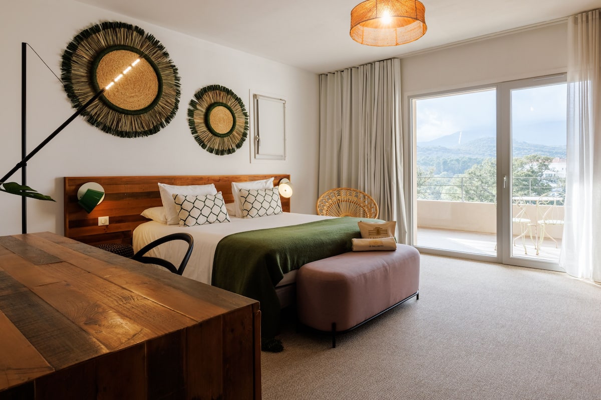 Quarto moderno com cama de madeira, manta verde e vista para o exterior através de grandes janelas.
