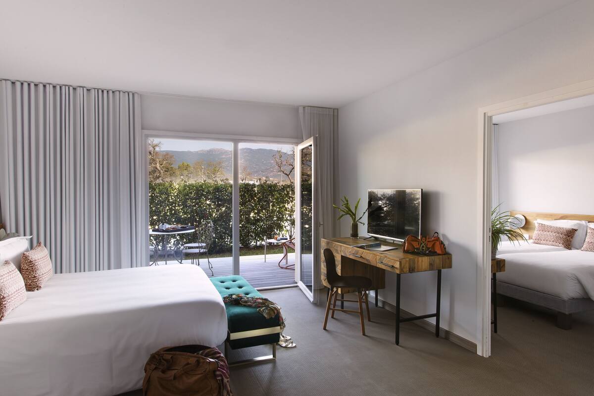 Quarto de hotel com cama, escrivaninha, cadeira, televisão e varanda com vista para árvores e montanhas.