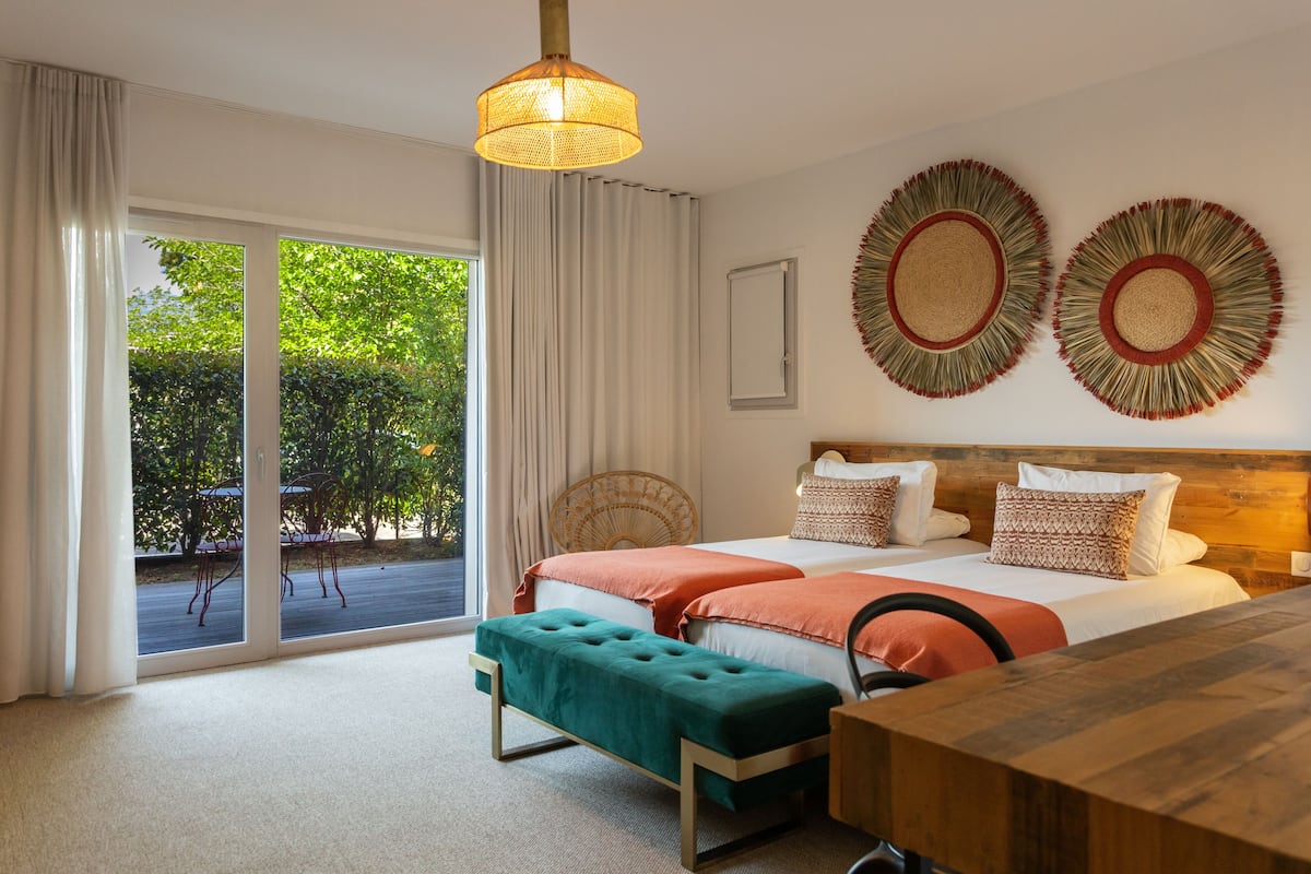 Quarto de hotel com uma grande porta de correr de vidro para o exterior, uma cama com duas almofadas, um banco e uma mesa de madeira.