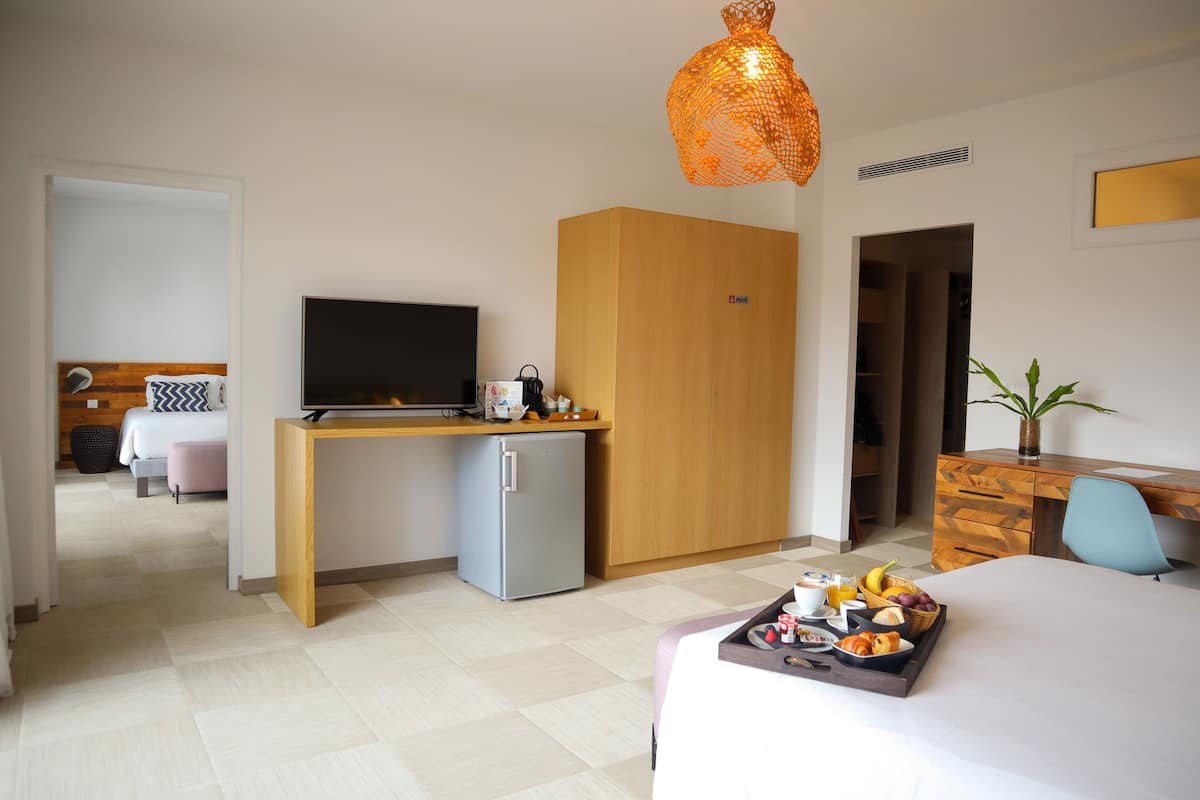Quarto de hotel moderno com cama, escrivaninha, televisão, mini-frigorífico e área de refeições com itens de café da manhã.