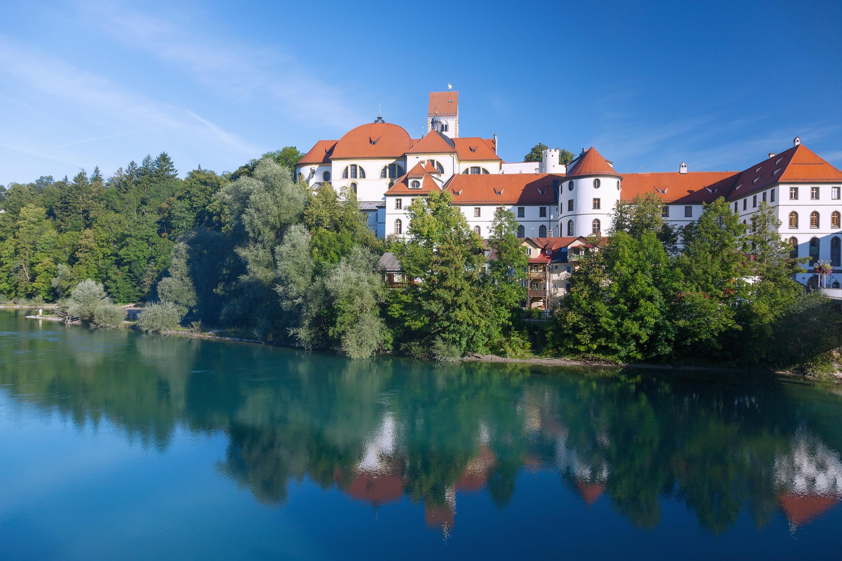Um edifício grande e histórico com várias estruturas de telhado vermelho, cercado por vegetação exuberante e refletido em águas calmas.