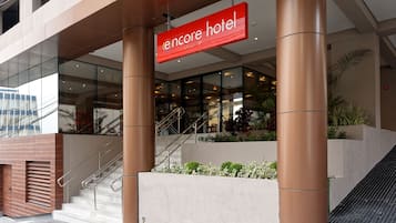 Entrada de hotel moderna com uma placa vermelha chamativa, grandes janelas de vidro e uma escadaria que leva ao saguão.