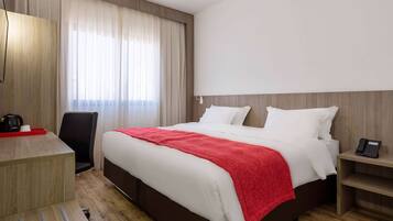 Quarto de hotel com cama, escrivaninha, cadeira, mesa de cabeceira com telefone e janela com cortinas.