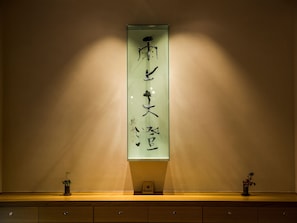Uma obra de caligrafia pendurada na parede, com caracteres chineses, acompanhada por duas pequenas plantas em vasos sobre uma prateleira de madeira.