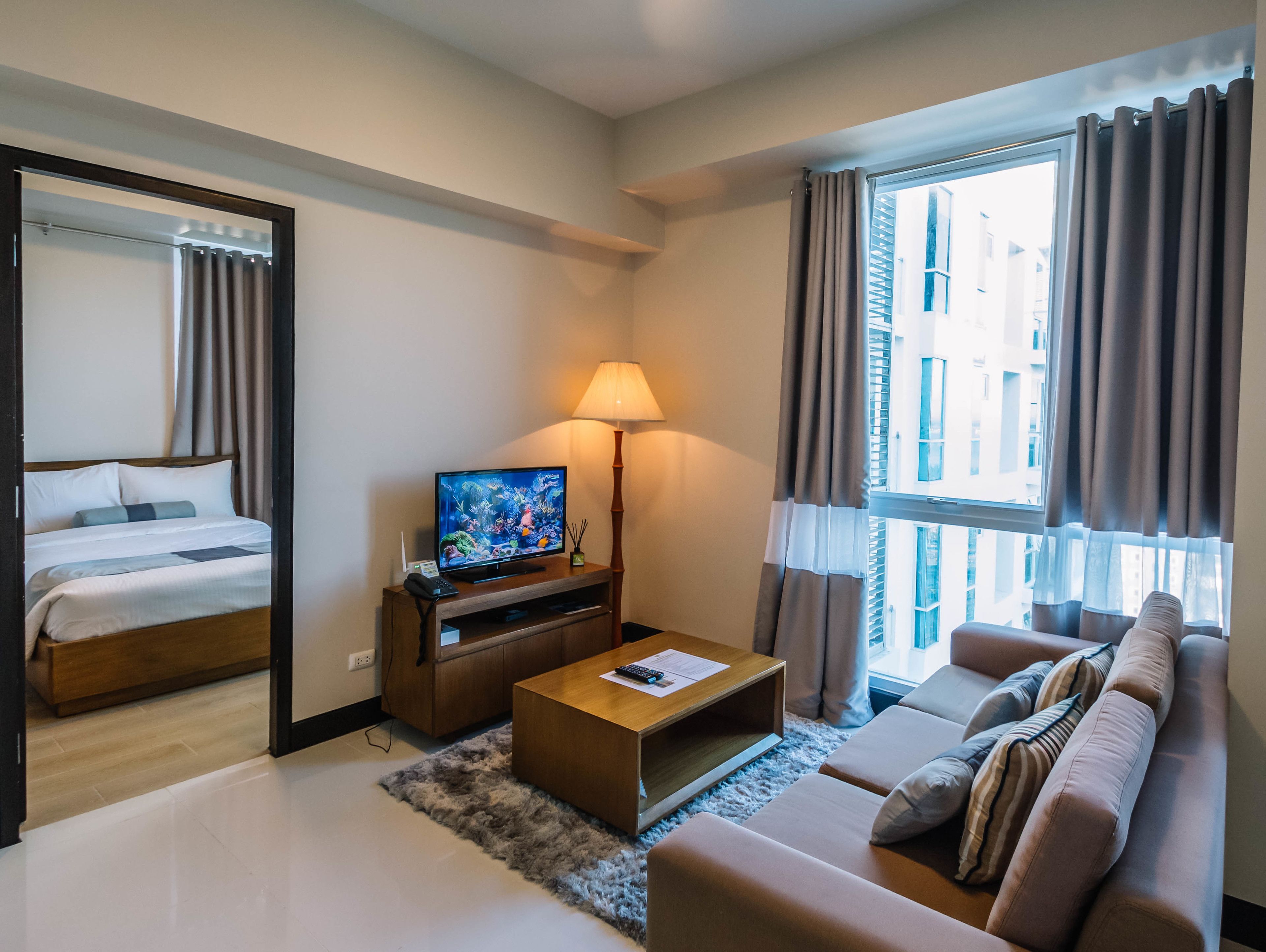 Quarto de hotel com cama, televisão sobre suporte de madeira, sofá, mesa de centro e luminária.